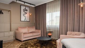 Quarto de hotel moderno com esquema de cores bege e rosa, apresentando um sofá, uma mesa redonda, uma cama e uma janela grande com cortinas.