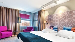 Quarto de hotel com uma cama grande, uma poltrona rosa, uma mesinha e uma janela com cortinas.
