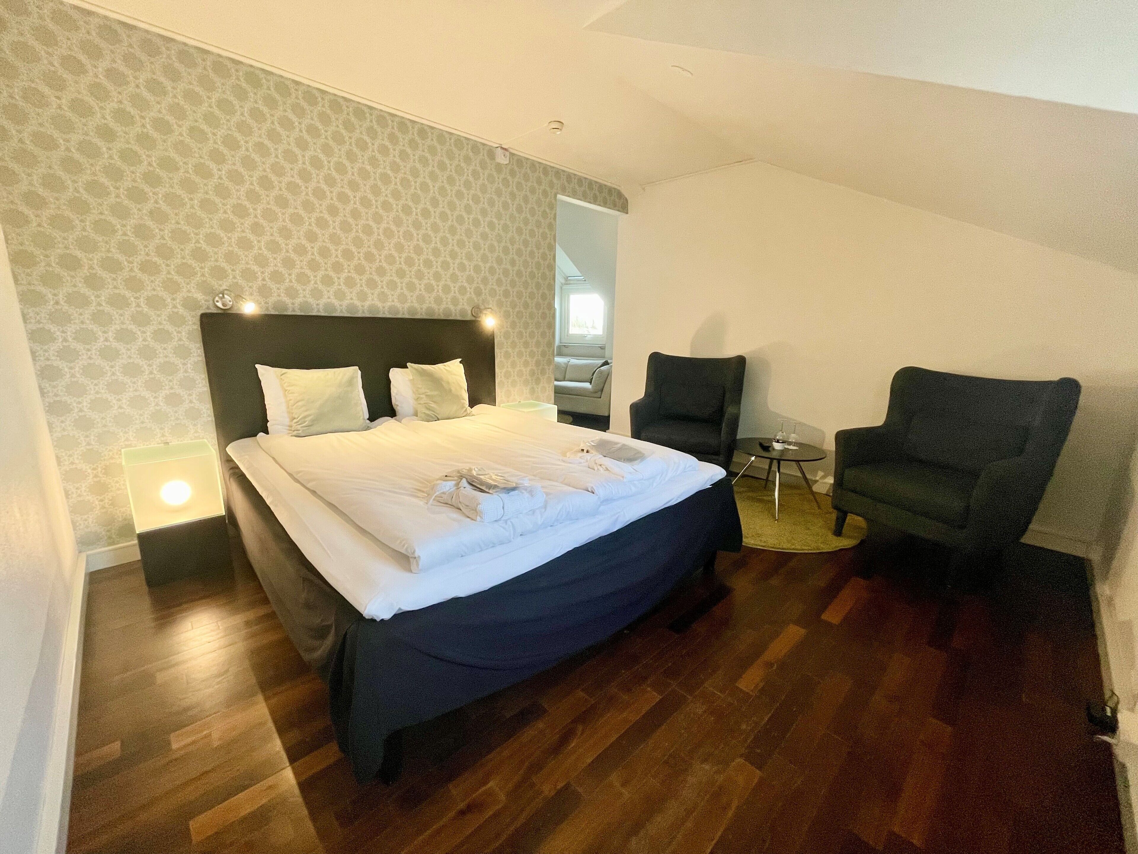 Quarto de hotel com uma cama grande, duas poltronas, uma mesinha e um espelho na parede.