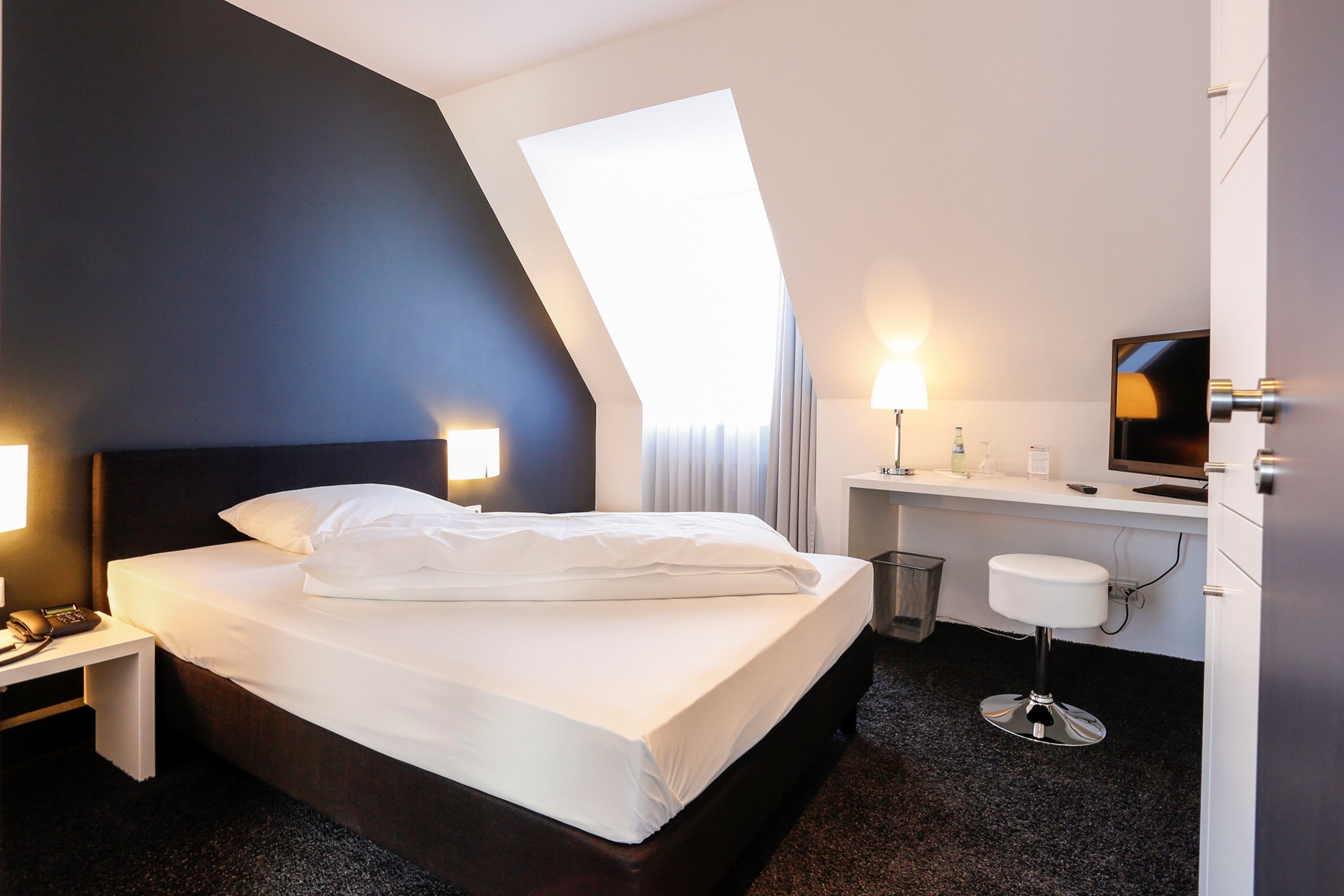 Quarto de hotel com cama, mesa de cabeceira, luminária e escrivaninha com cadeira.
