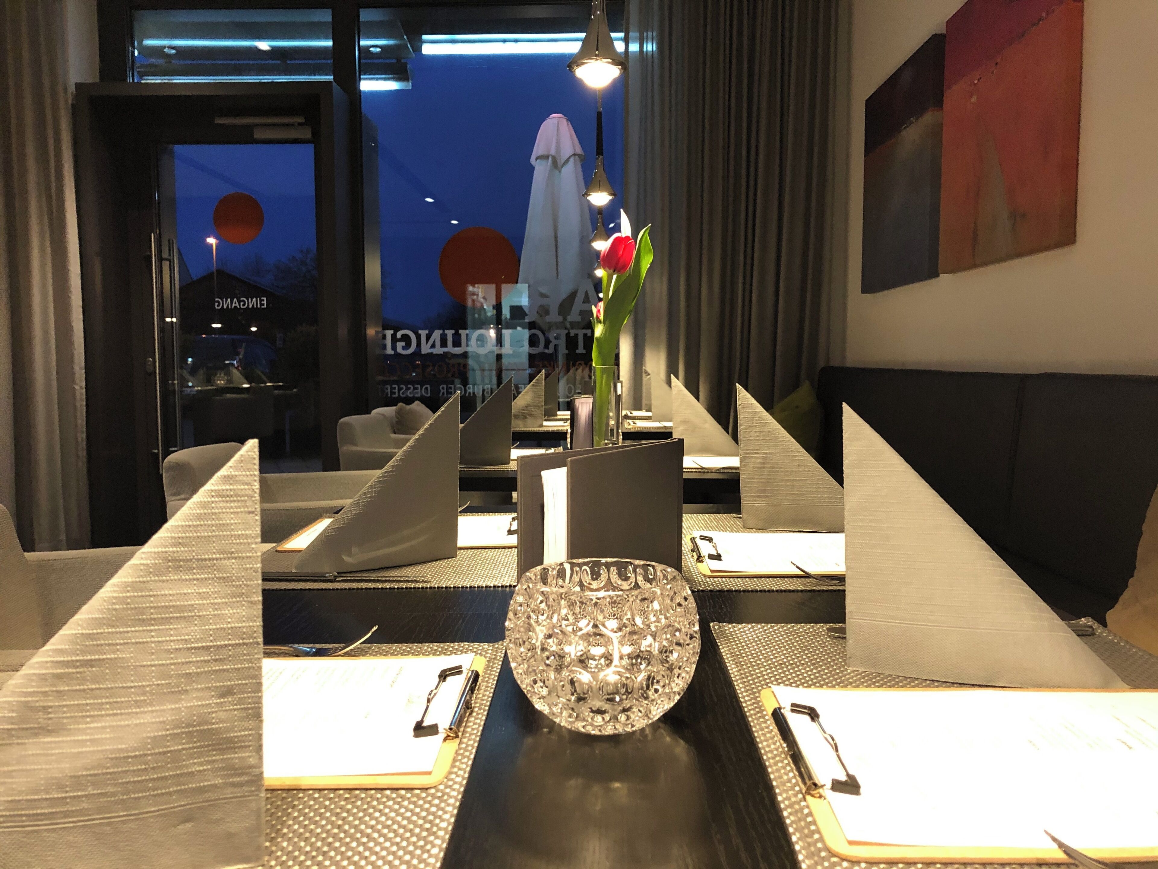 Quarto de hotel moderno com uma mesa de jantar comprida, um vaso com flores e vista para a paisagem urbana pela janela.
