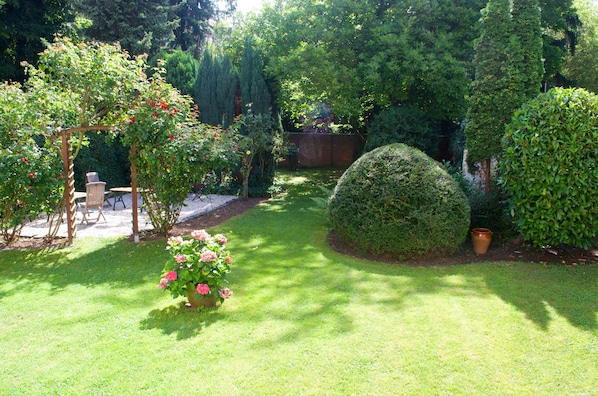 Um jardim bem cuidado com uma pérgola, uma mesa redonda e cadeiras, cercado por arbustos variados e um sebedeio bem aparado.