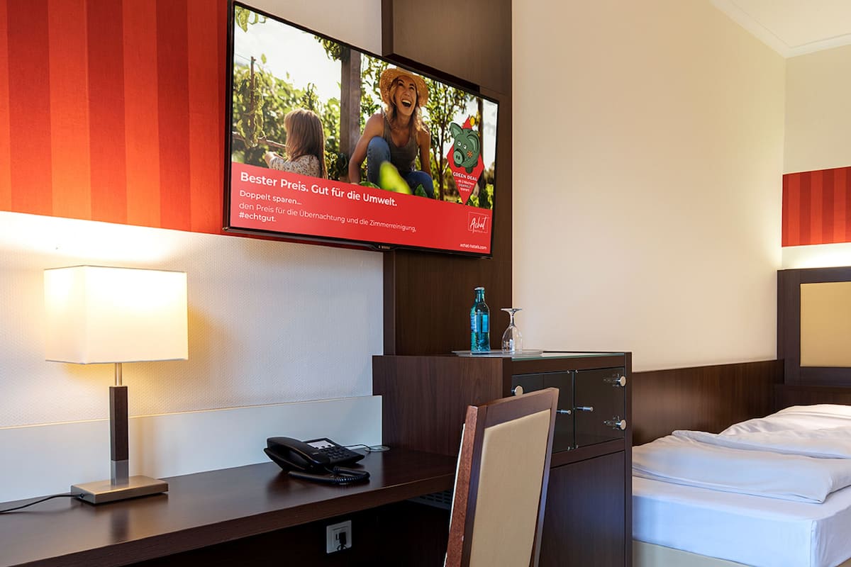 Quarto de hotel com cama, escrivaninha com luminária, televisão na parede e espelho sobre a cama.