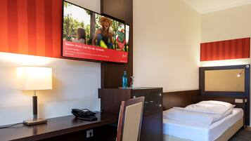 Quarto de hotel com cama, escrivaninha com luminária, televisão na parede e espelho sobre a cama.