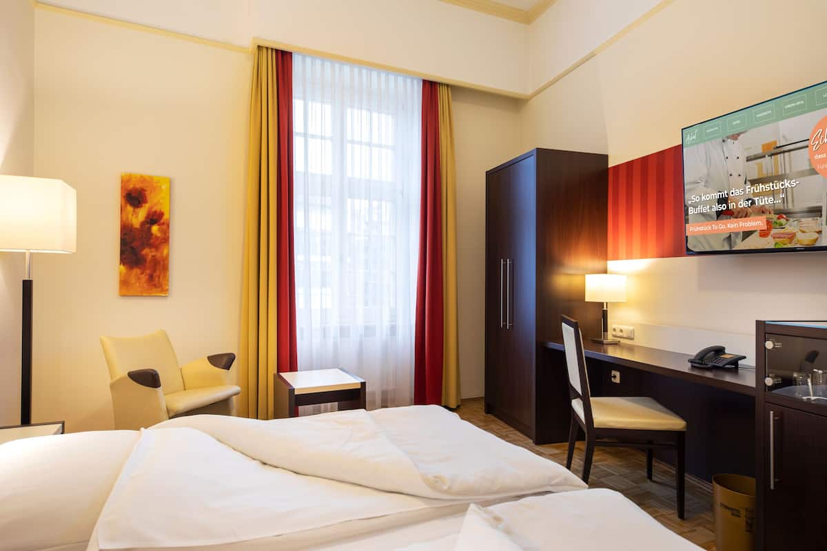 Quarto de hotel com cama, escrivaninha, cadeira, televisão e janela com cortinas.