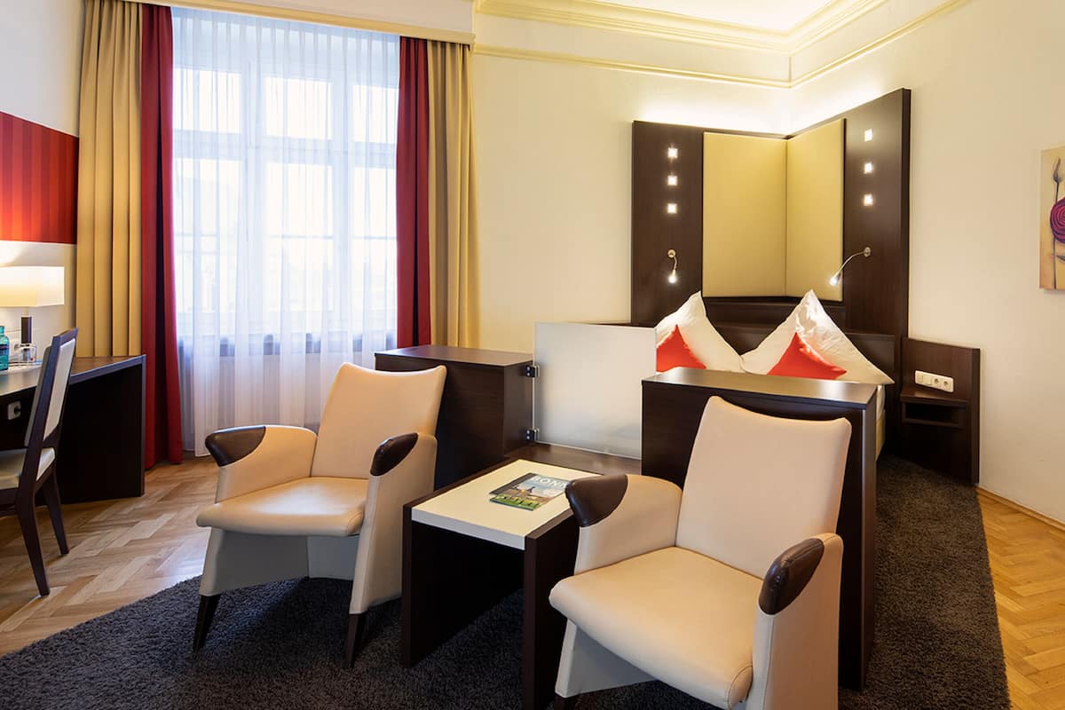 Quarto de hotel com duas poltronas, uma mesinha, uma escrivaninha, uma luminária, um espelho e um quadro na parede.