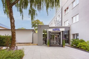 Entrada de um hotel moderno com porta de vidro, placa de identificação e plantas em vasos.