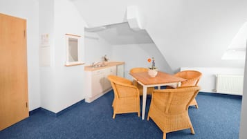 Um quarto pequeno e iluminado, com uma cozinheneta, uma mesa de jantar e cadeiras de vime, e um tapete azul.