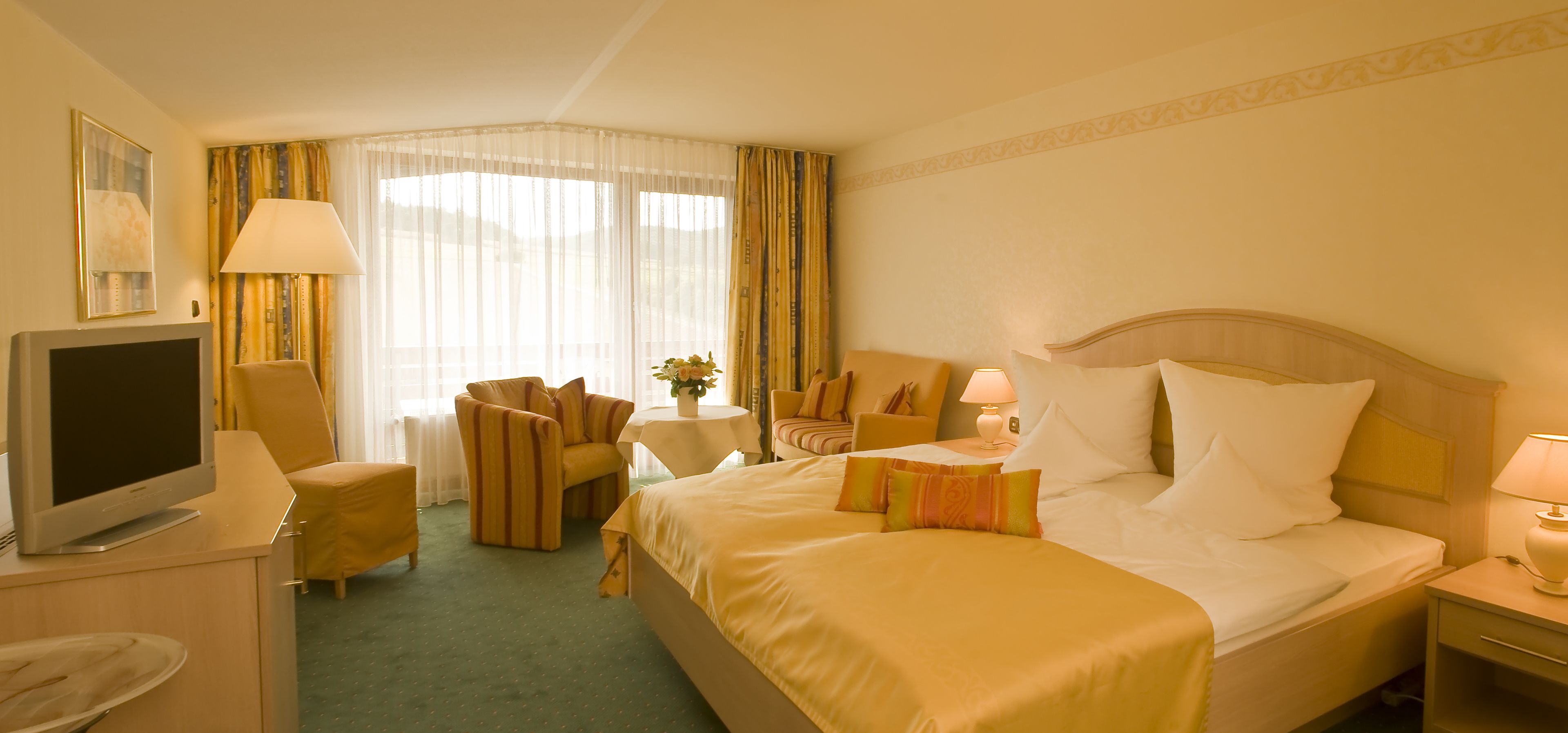 Quarto de hotel com cama, televisão, escrivaninha, cadeira, mesinha de centro e cortinas.