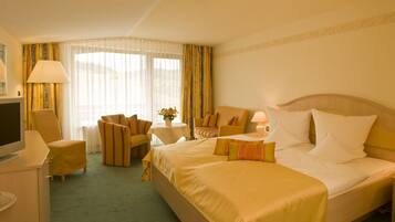 Quarto de hotel com cama, televisão, escrivaninha, cadeira, mesinha de centro e cortinas.