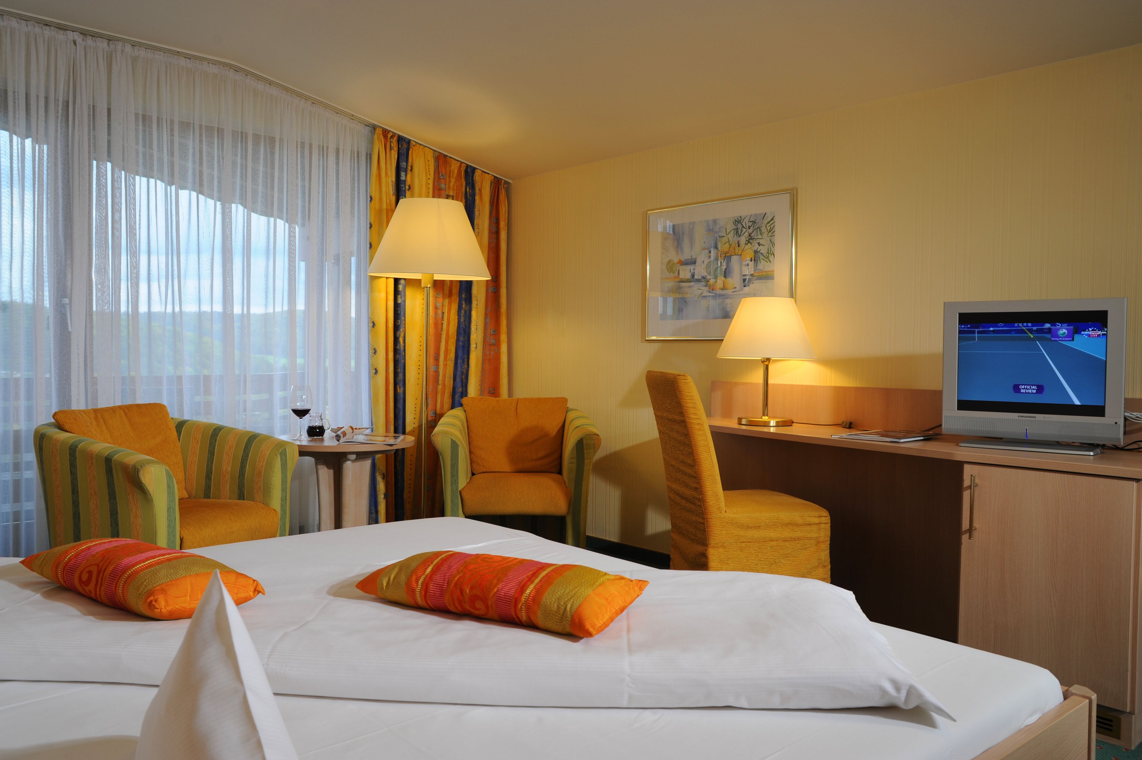 Quarto de hotel com uma cama, duas poltronas, uma escrivaninha com televisão e um quadro na parede.