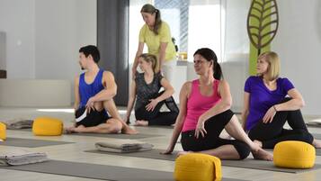 Um grupo de pessoas numa aula de yoga, sentadas em tapetes com almofadas amarelas, num espaço com uma grande decoração em forma de folha na parede.