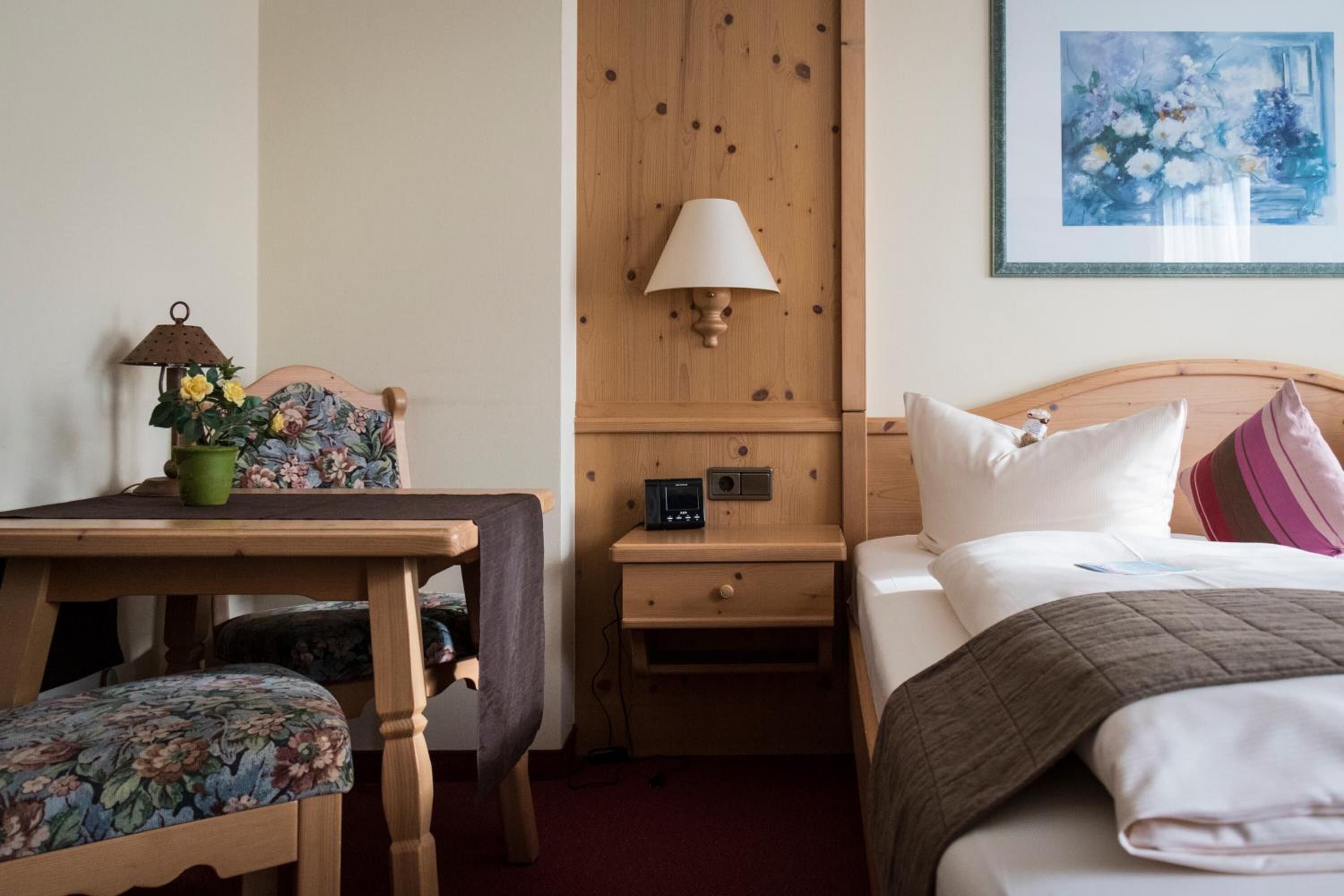 Quarto de hotel com cabeceira de madeira, mesa de cabeceira com abajur, rádio, poltrona florida e cama com almofadas.