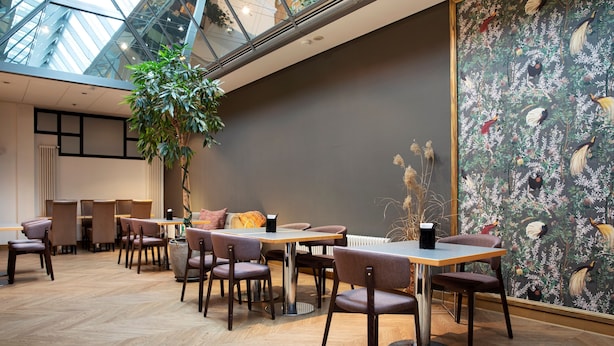 Um interior moderno com teto alto, área de estar central com mesas e cadeiras, uma planta grande e uma parede decorativa com um mural de tema natural.