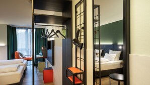 Quarto de hotel com duas camas, uma escrivaninha, uma cadeira, um espelho e um cabideiro.