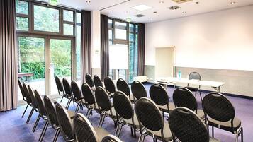 Uma sala de conferências com fileiras de cadeiras, grandes janelas, uma mesa e uma tela de projeção.