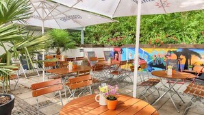 Um café ao ar livre com mesas e cadeiras de madeira, guarda-sóis e um mural na parede.