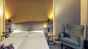Quarto de hotel com uma cama grande, cabeceira estampada, duas luminárias de cabeceira, uma cadeira e uma mala.