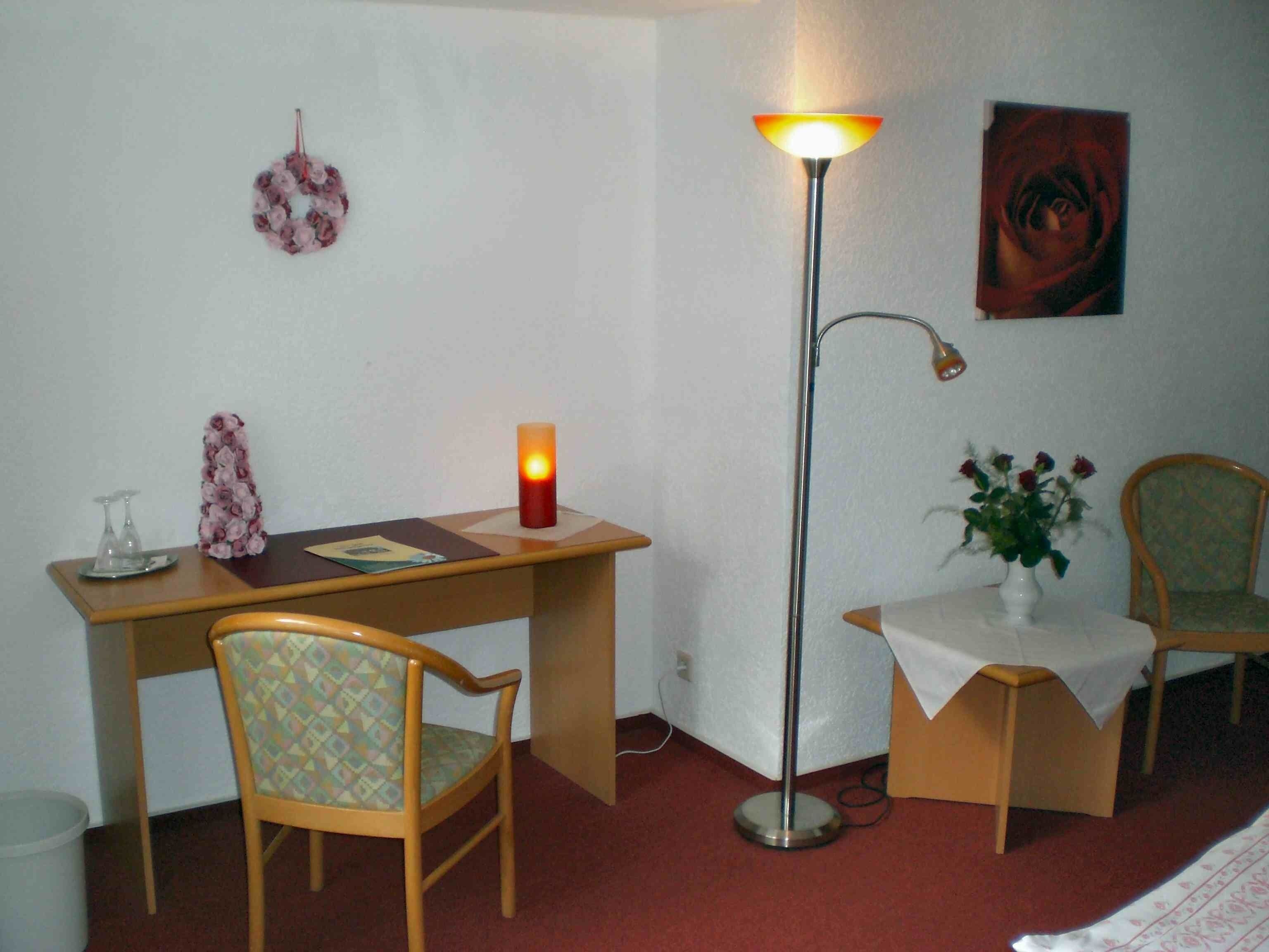 Quarto pequeno e bem arrumado com escrivaninha, duas cadeiras, abajur, vela, um vaso com flores e um quadro na parede.