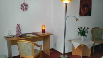 Quarto pequeno e bem arrumado com escrivaninha, duas cadeiras, abajur, vela, um vaso com flores e um quadro na parede.
