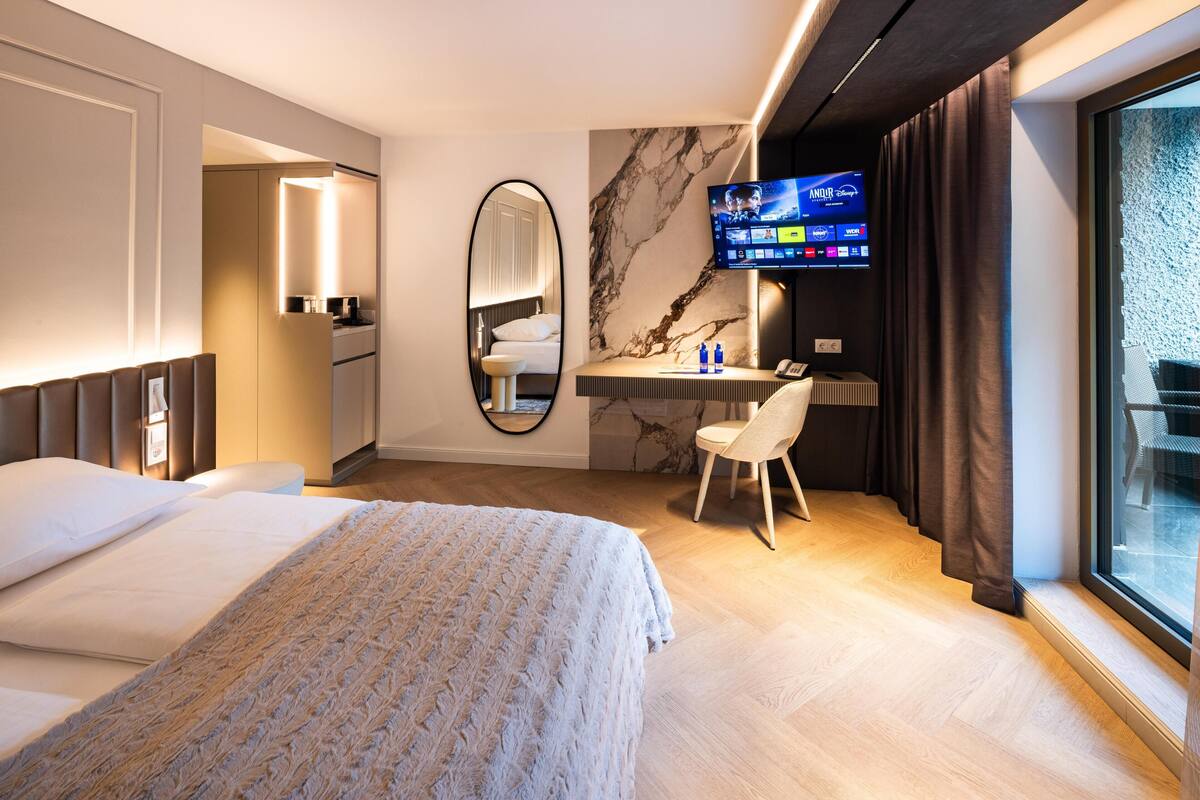 Quarto de hotel moderno com uma cama grande, uma escrivaninha com cadeira, televisão na parede e uma janela ampla com vista.