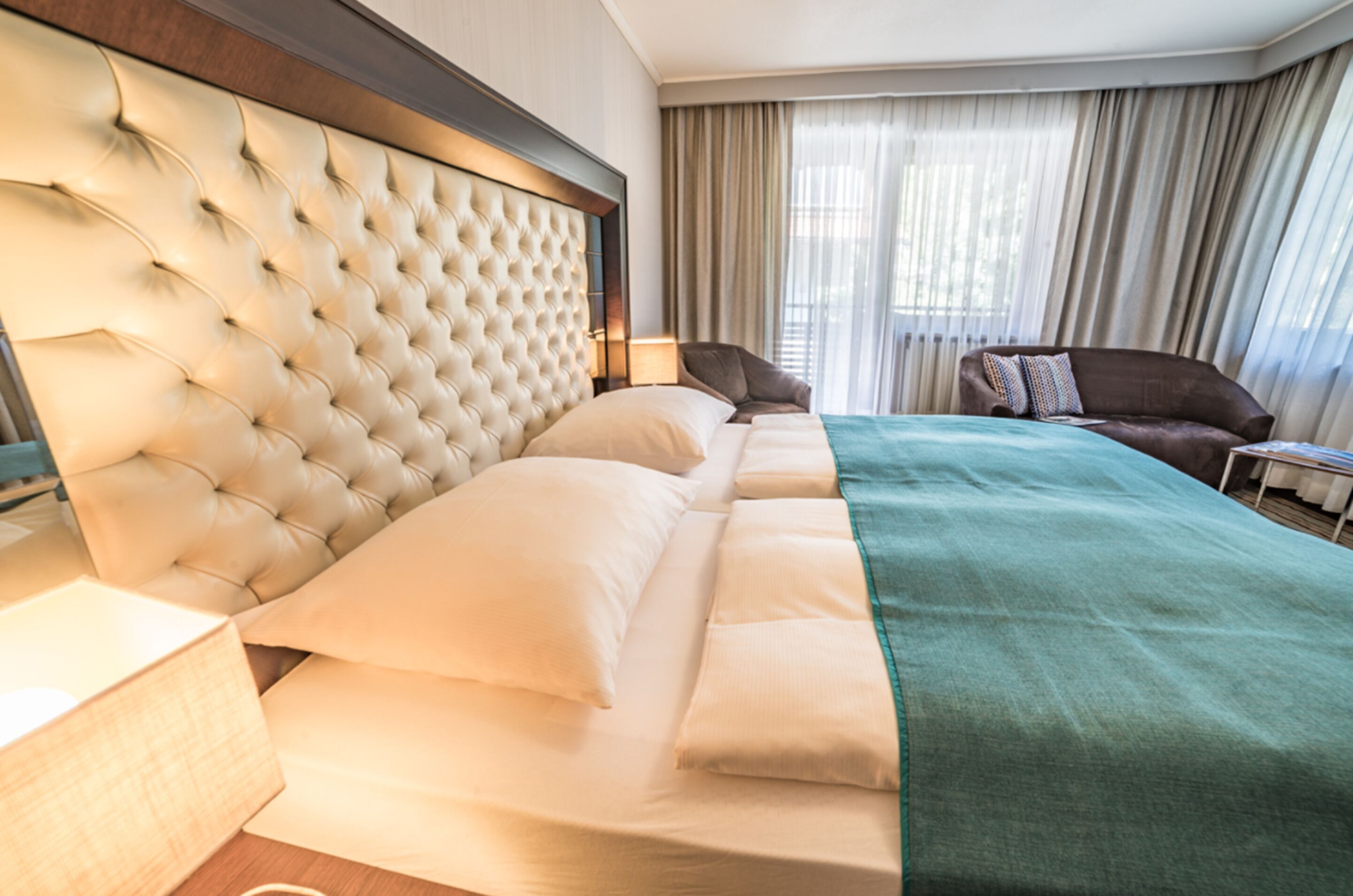 Quarto de hotel com uma cama grande, cabeceira estofada, um sofá e vista para o exterior através de grandes janelas.