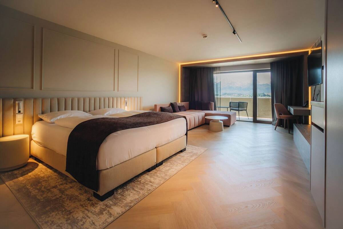 Quarto de hotel moderno com uma cama grande, um sofá, uma mesa pequena e uma varanda com vista.