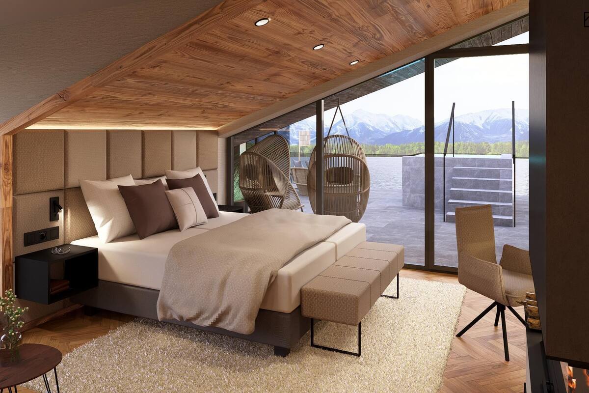 Quarto moderno com uma cama grande, mesas de cabeceira, um espaço de estar com cadeiras e vista para montanhas através de amplas janelas.