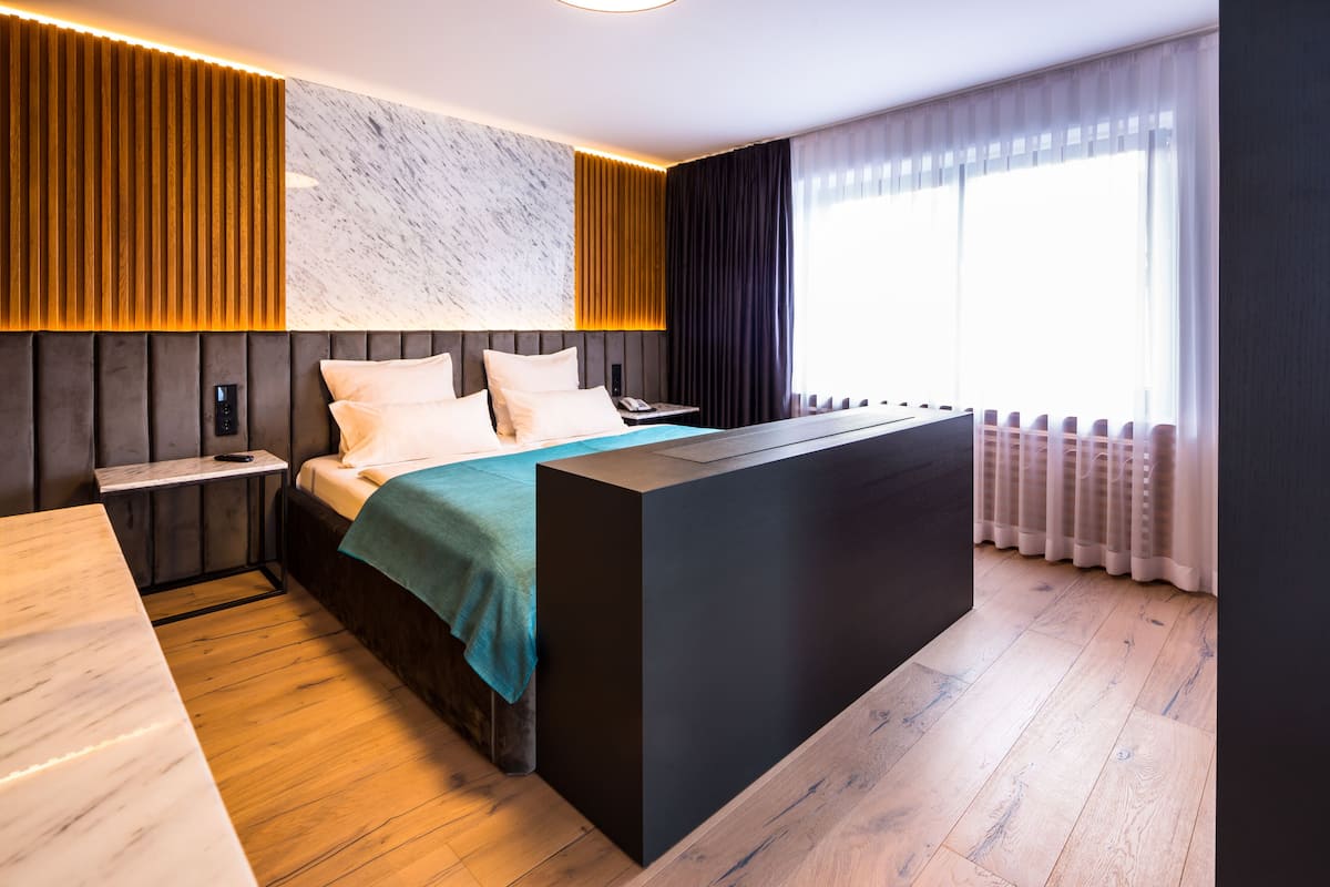 Quarto de hotel moderno com uma cama grande, uma mesinha de cabeceira, escrivaninha embutida e uma janela ampla com cortinas.