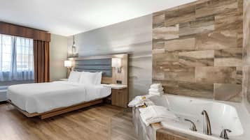 Quarto de hotel com cama, mesa de cabeceira, banheiro com banheira e uma janela grande com cortinas.