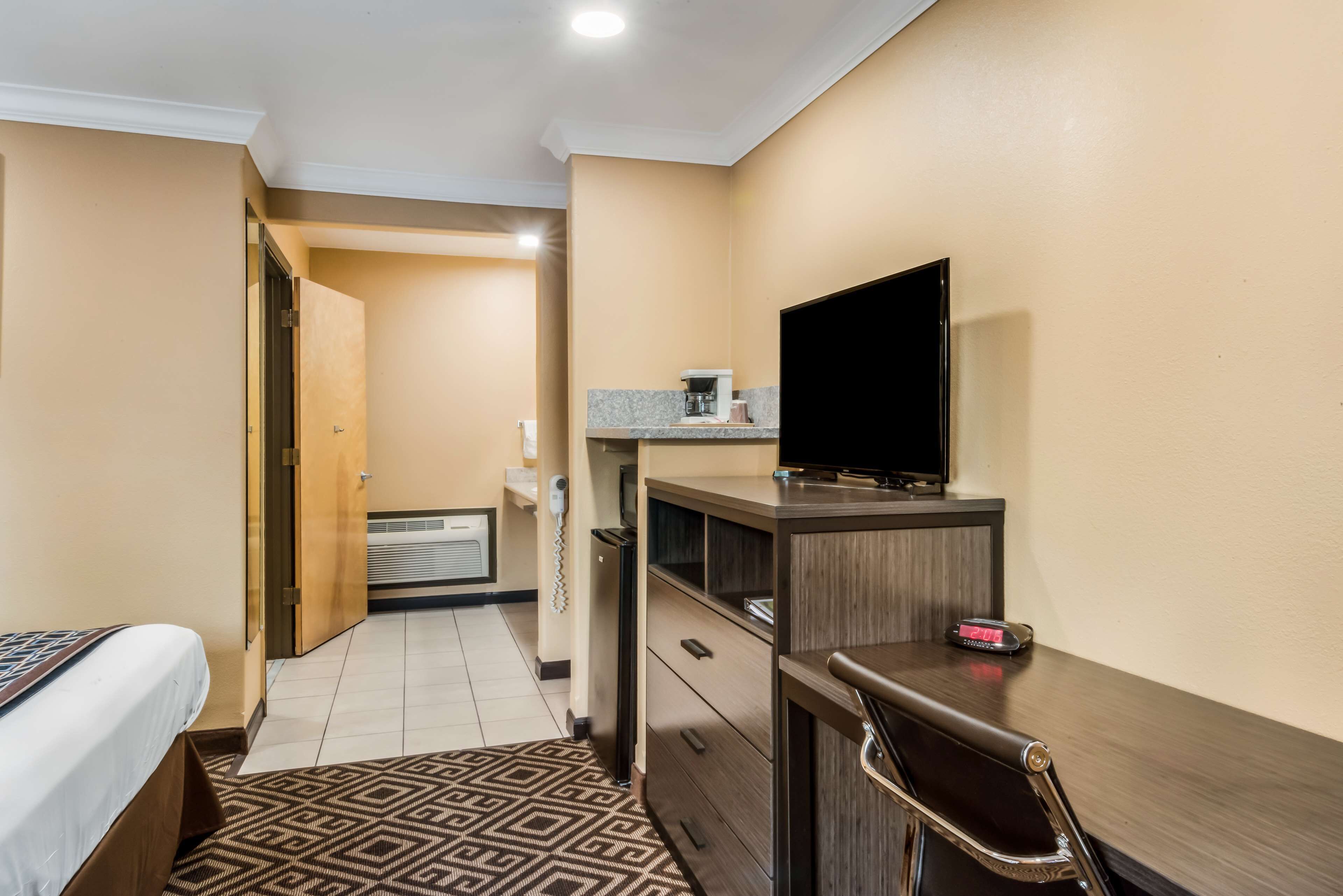 Quarto de hotel com cama, secretária com televisão de ecrã plano, uma cadeira, kitchenette pequena e tapete estampado.