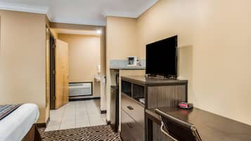 Quarto de hotel com cama, secretária com televisão de ecrã plano, uma cadeira, kitchenette pequena e tapete estampado.