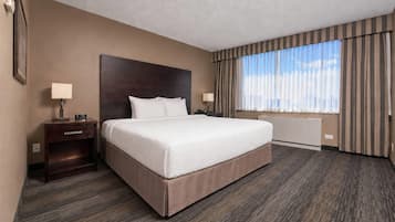 Quarto de hotel com uma cama grande, uma mesa de cabeceira, um abajur, uma janela com cortinas e um radiador.