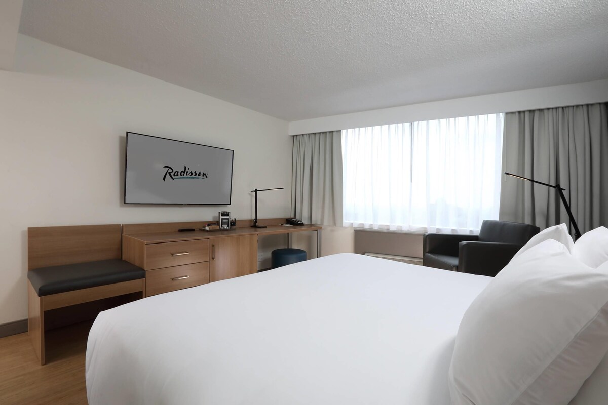 Quarto de hotel com cama grande, escrivaninha com cafeteira, cadeira e televisão fixada na parede.