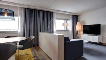 Quarto de hotel moderno com área de refeições, sofá, TV e uma janela grande com cortinas.