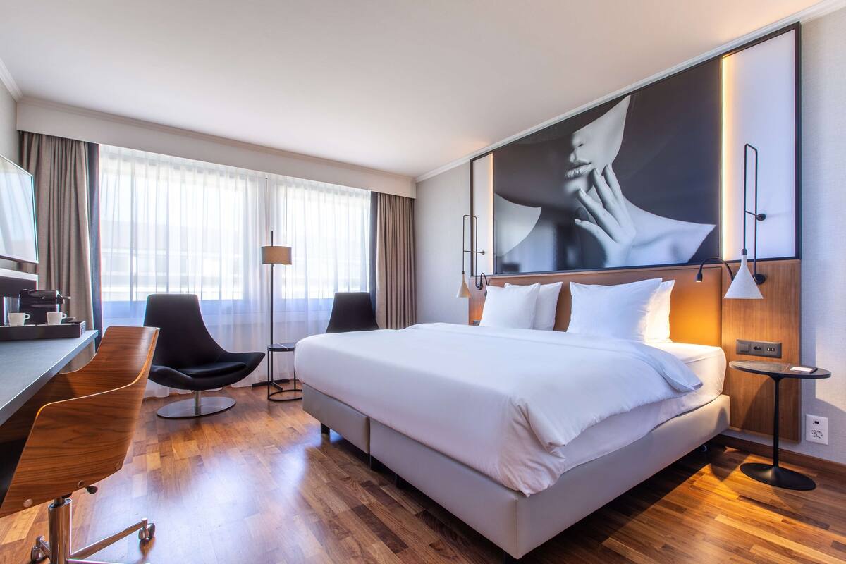 Quarto de hotel moderno com uma cama grande, uma escrivaninha com cadeira, televisão de tela plana e uma pintura abstrata grande na parede.