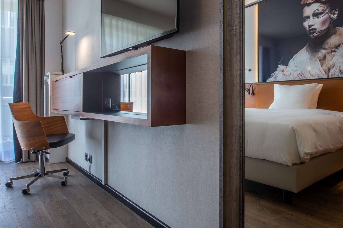 Quarto de hotel moderno com televisão de tela plana na parede, escrivaninha de madeira com cadeira, cama com roupa de cama branca e um espelho grande refletindo o quarto.