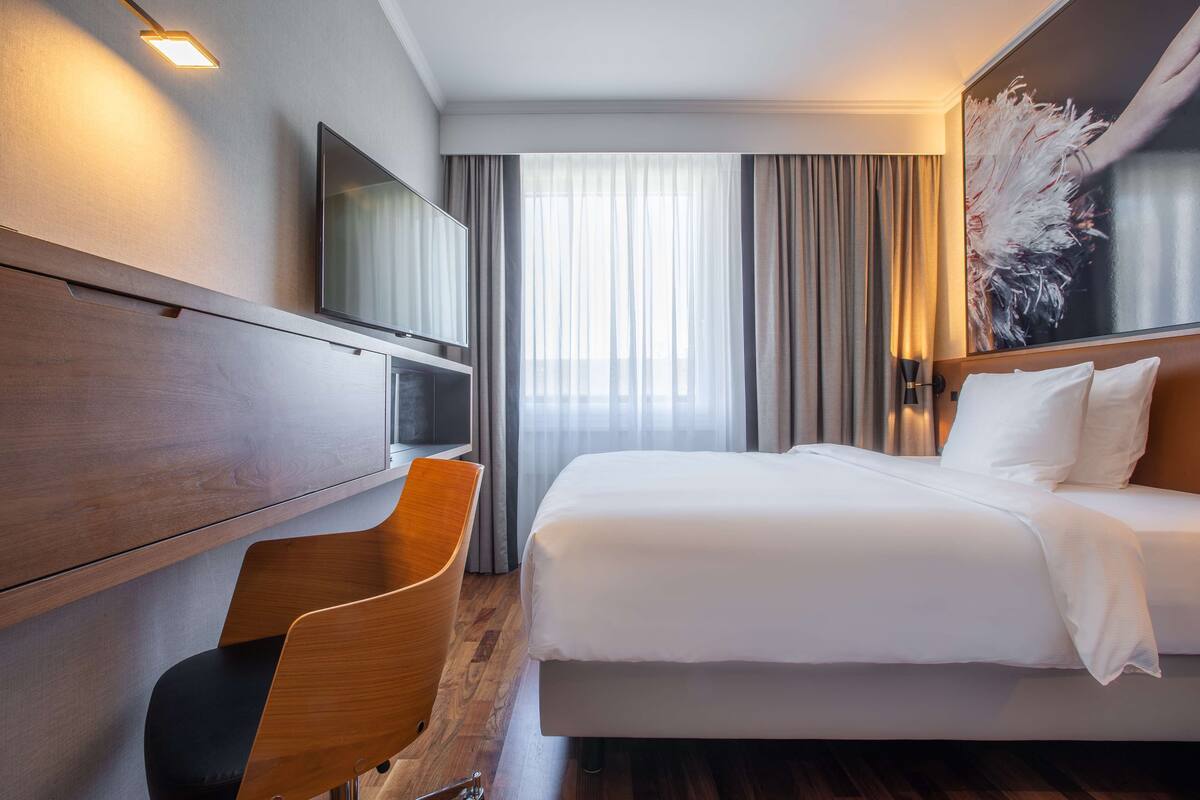 Quarto de hotel moderno com uma cama grande, televisão fixada na parede, uma cadeira de madeira e uma janela ampla com cortinas.