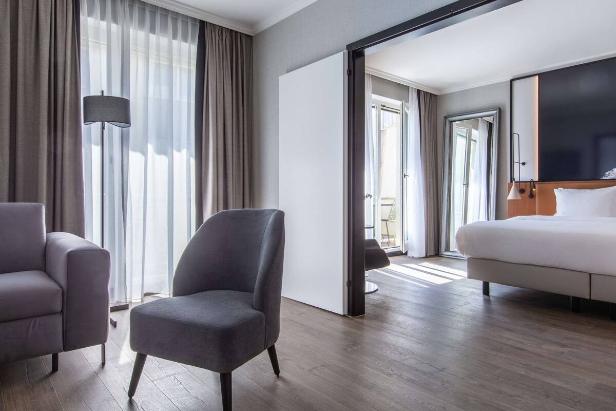 Quarto de hotel moderno com uma cama grande, área de estar com duas poltronas e vista através de uma porta de vidro.
