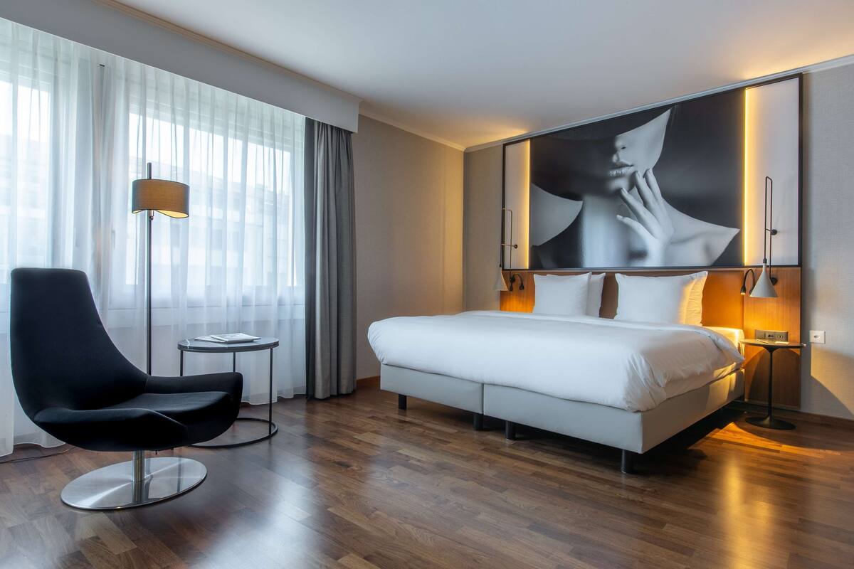 Quarto de hotel moderno com uma cama grande, uma cadeira preta, uma mesa pequena e uma grande obra de arte na parede.