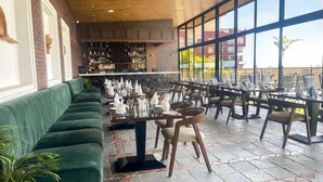 Restaurante com um sofá verde, mesas de jantar com guardanapos e pratos brancos, e área de bar com prateleiras cheias de garrafas.