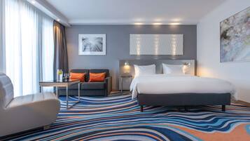 Quarto de hotel com uma cama grande, um sofá, uma pequena mesa com copos e vista para o exterior através de janelas amplas.