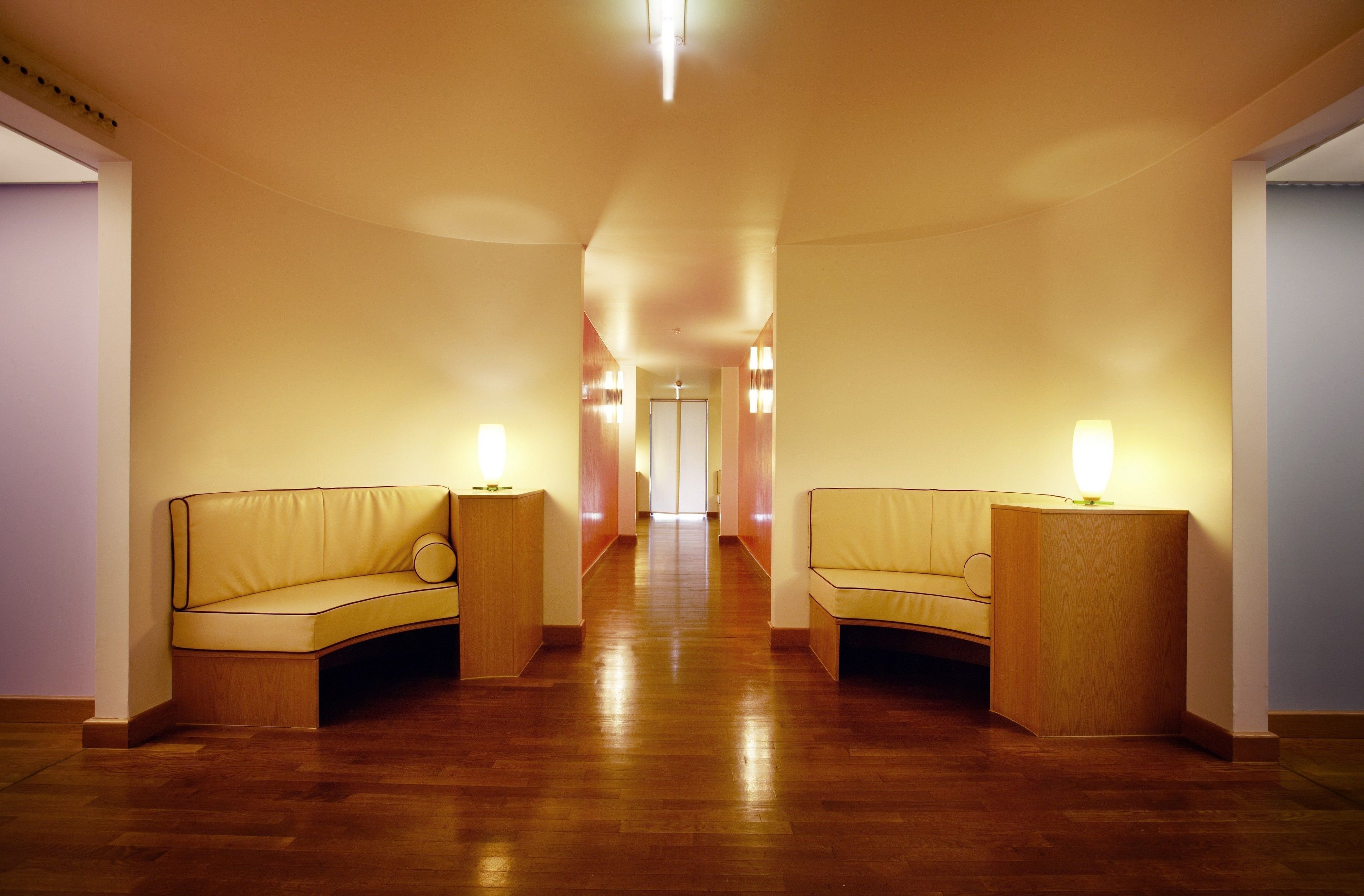 Corredor de hotel com dois sofás e luminárias, piso de madeira e vista para um quarto no final.