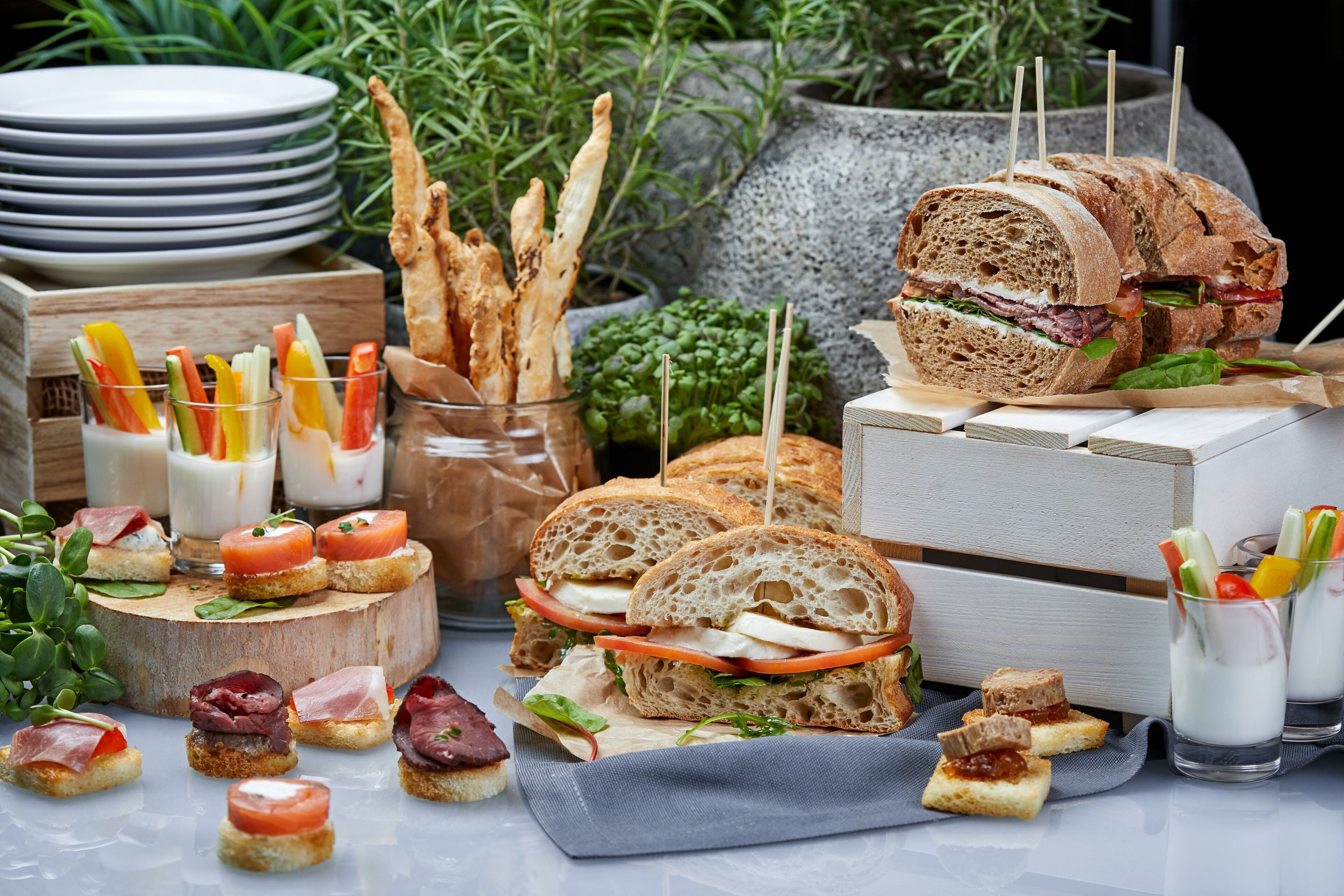 Diversos alimentos, incluindo sanduíches, aperitivos e bebidas, dispostos sobre uma mesa com fundo de madeira rústica.