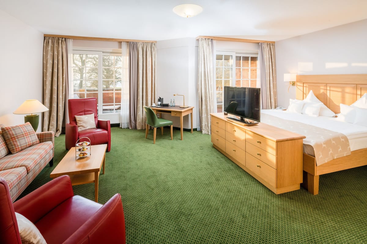 Quarto de hotel com cama, escrivaninha, televisão, poltrona vermelha, sofá e tapete verde.
