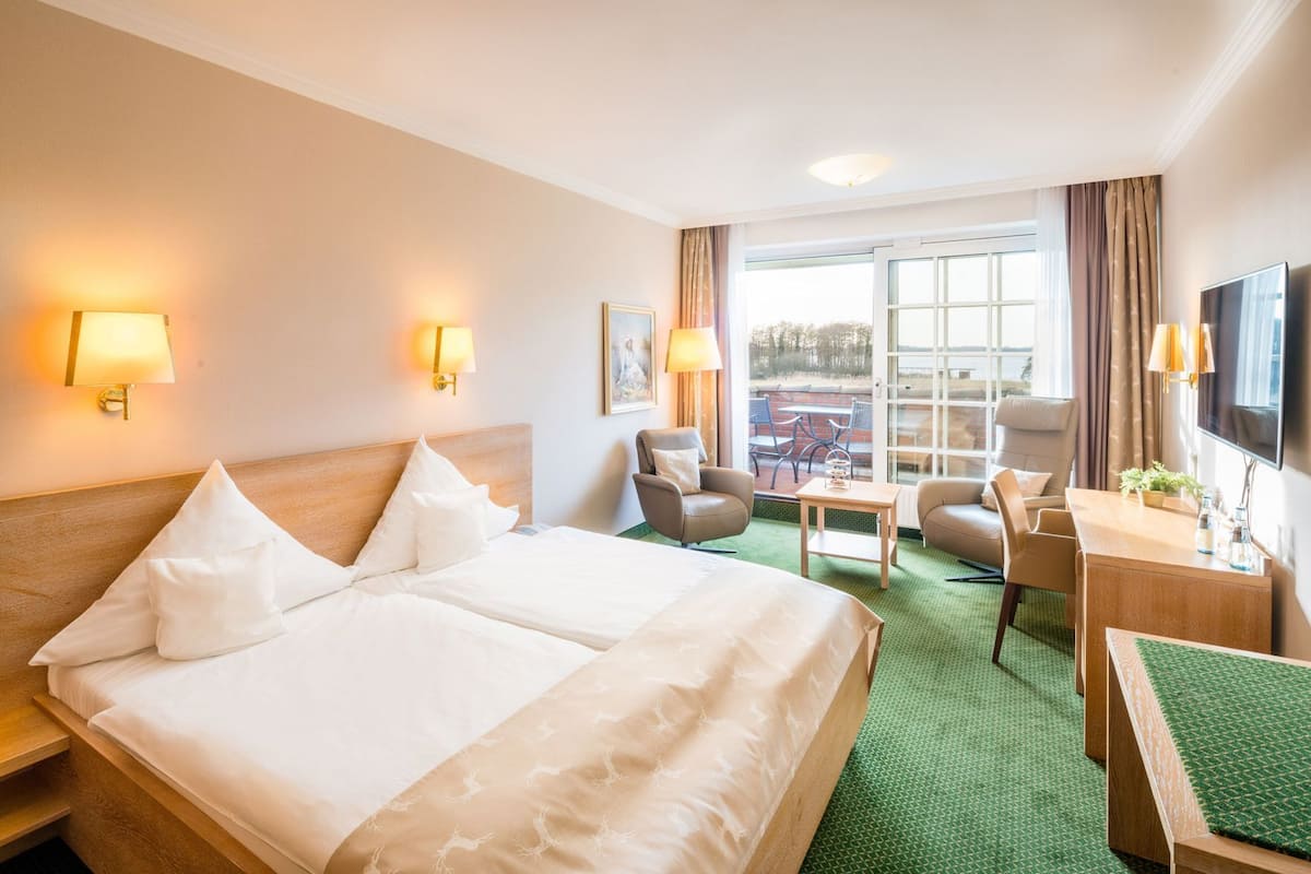 Quarto de hotel com uma cama grande, duas poltronas, uma mesa pequena, escrivaninha, televisão e janela com vista.