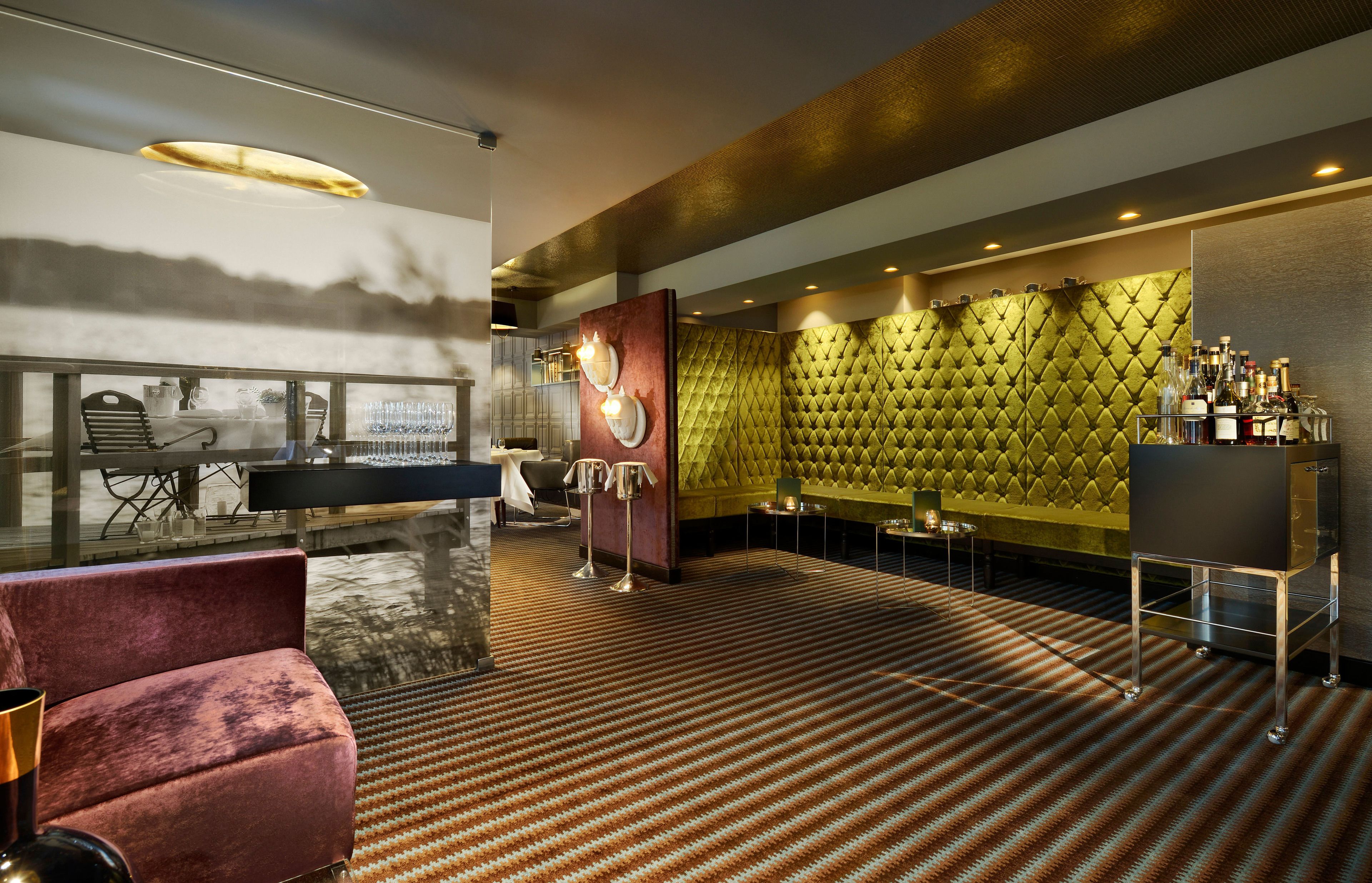 Uma área de bar moderna com um painel verde texturizado, um balcão com garrafas e um espaço de estar com uma cadeira vermelha e uma mesa.