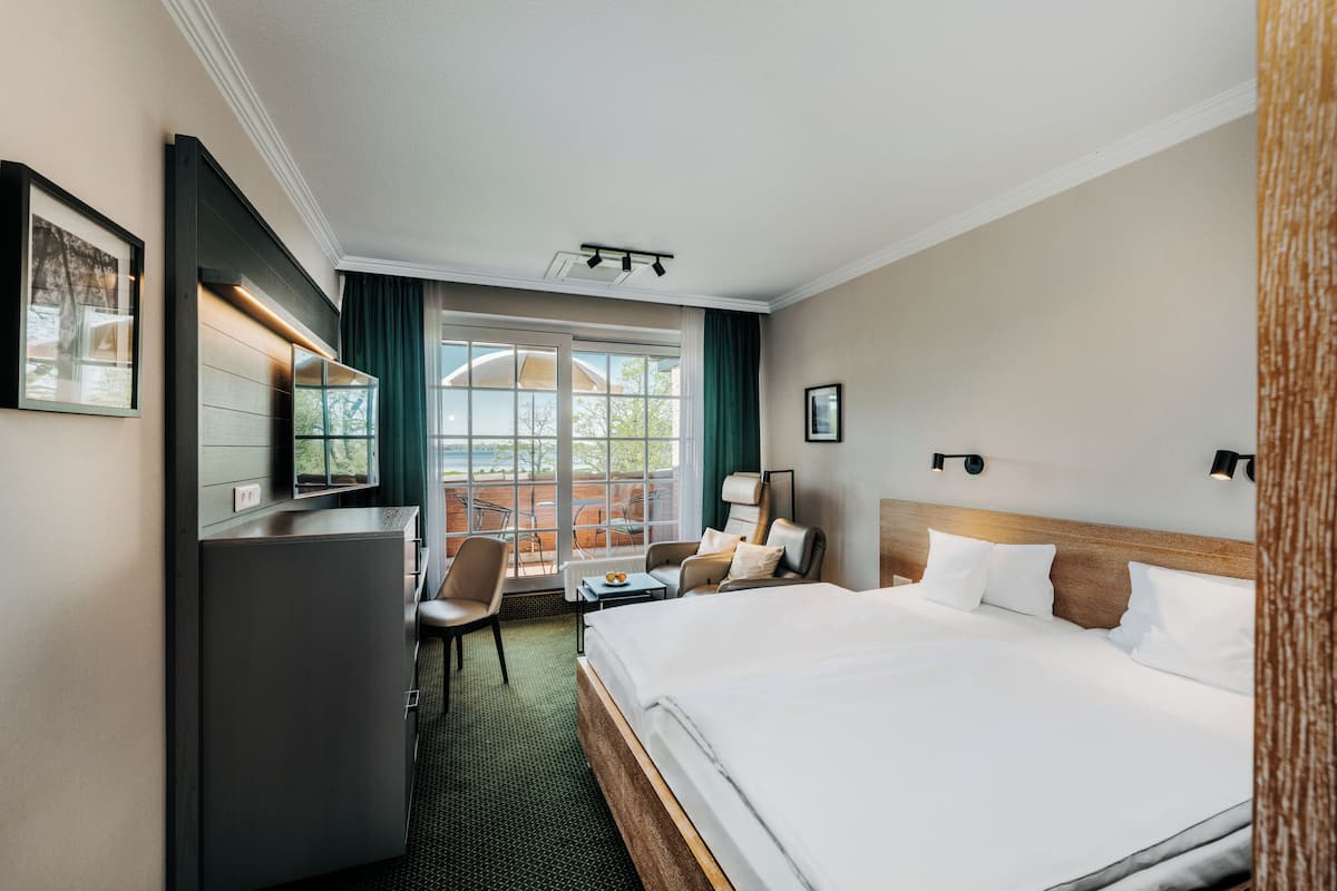 Quarto de hotel com uma cama grande, uma mesa, uma cadeira e vista para o exterior pela janela.