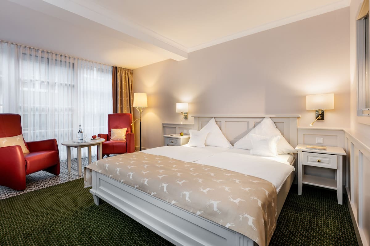 Quarto de hotel com uma cama grande, duas mesas de cabeceira, uma poltrona vermelha, uma mesinha com abajur e uma janela com cortinas leves.
