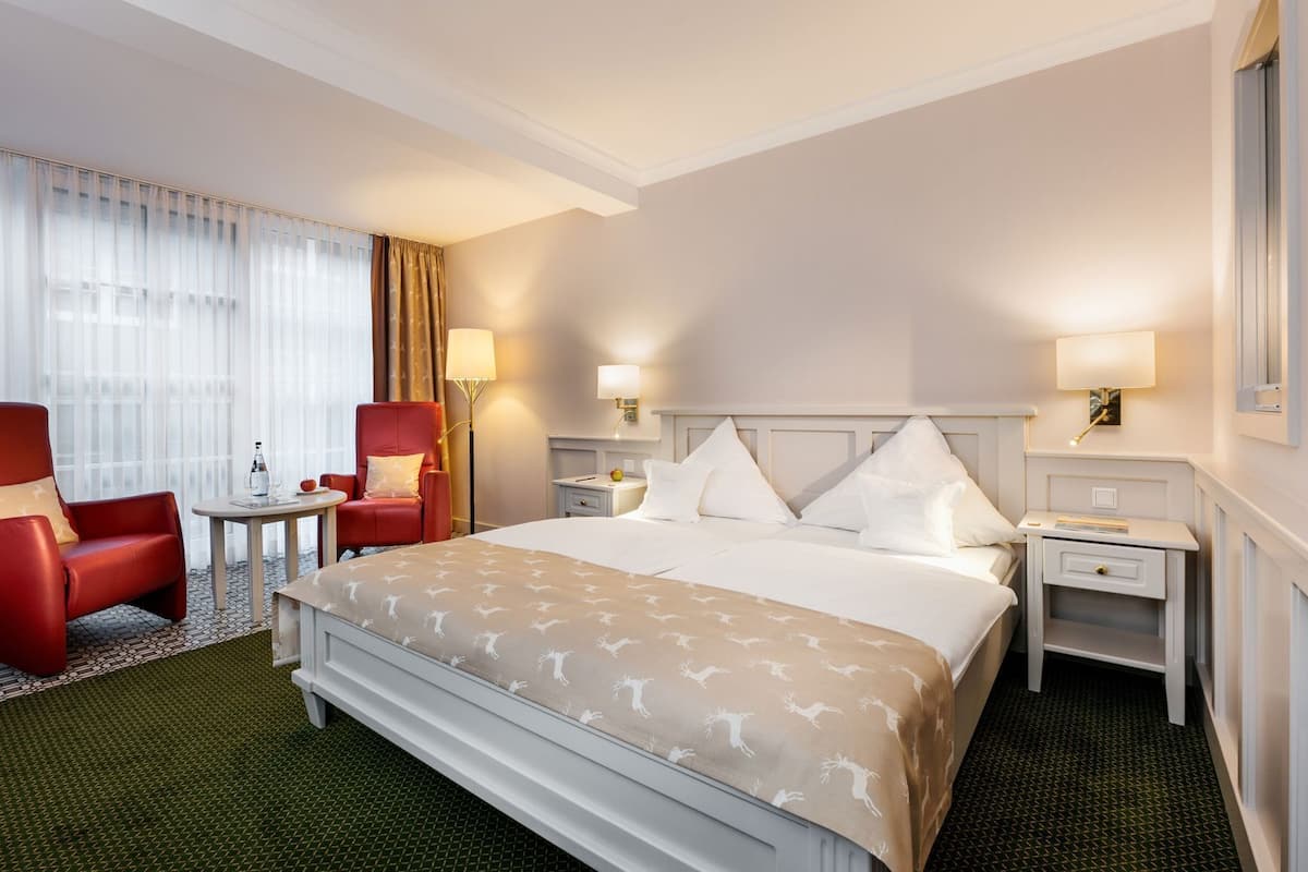 Quarto de hotel com uma cama grande, uma poltrona vermelha, uma mesinha de cabeceira com abajur e uma janela com cortinas.